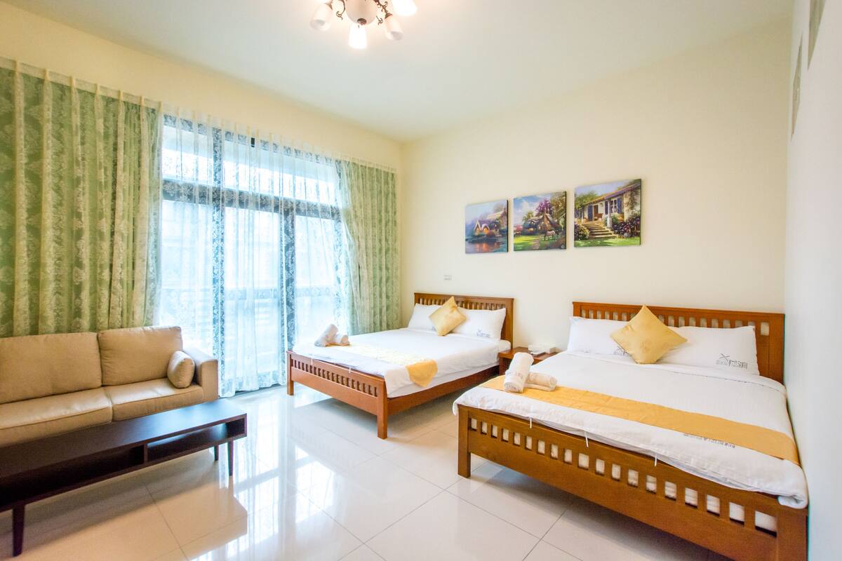 Quarto de hotel com duas camas de madeira, um sofá bege e uma janela grande com cortinas leves.