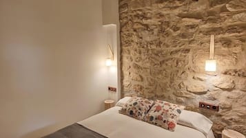 Quarto com parede de pedra, vigas de madeira, cama com almofadas florais e duas luzes fixadas na parede.