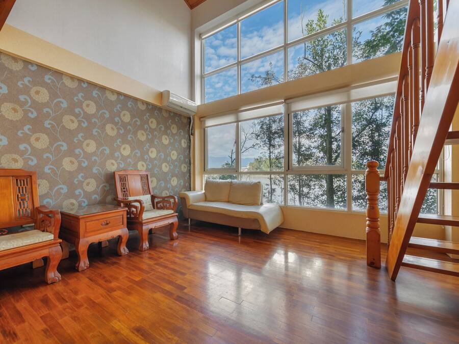 Um quarto espaçoso com piso de madeira, um sofá, um armário de madeira e uma grande janela com vista para árvores.