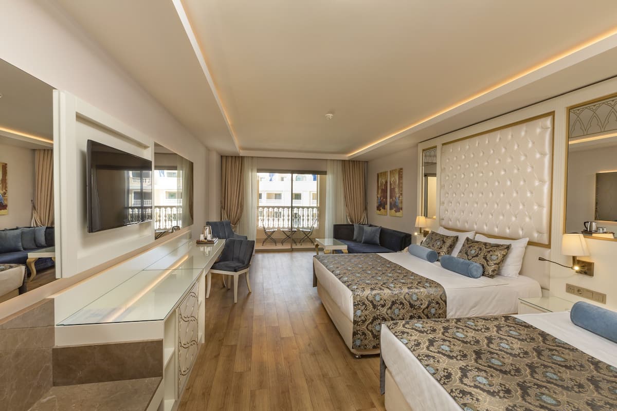 Quarto de hotel com uma cama grande, uma cama menor, escrivaninha, cadeira, televisão e varanda com cortinas.