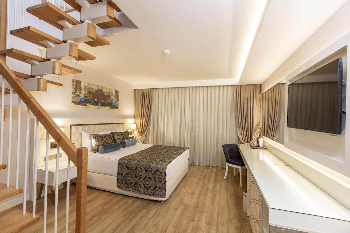 Quarto de hotel moderno com escada de madeira, cama grande com roupa de cama estampada, escrivaninha com cadeira e uma janela ampla com cortinas.