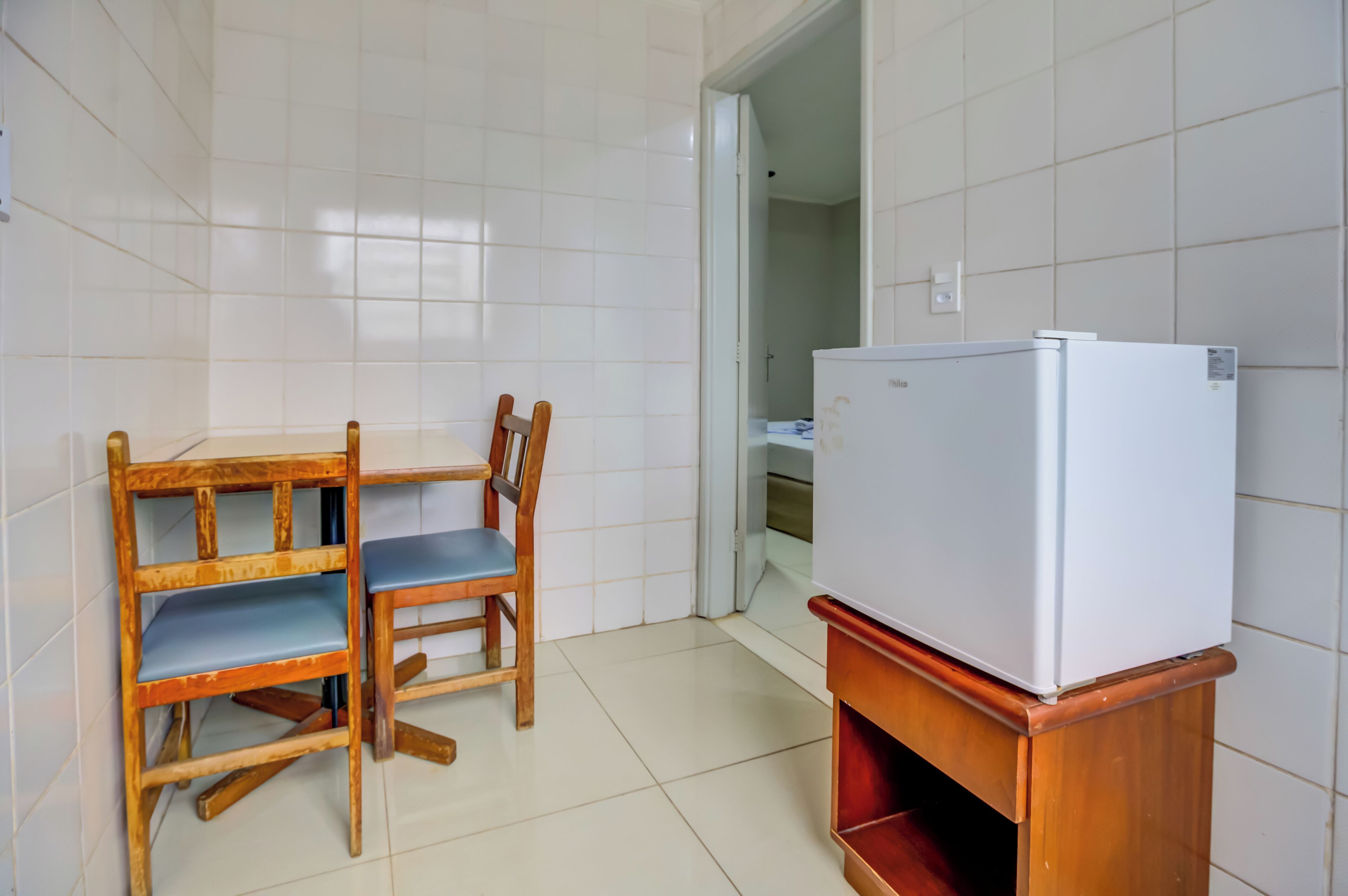 Uma pequena cozinha compacta com geladeira branca, mesa de madeira e cadeiras azuis, e paredes com azulejos brancos.