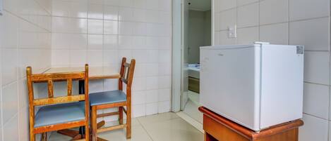 Uma pequena cozinha compacta com geladeira branca, mesa de madeira e cadeiras azuis, e paredes com azulejos brancos.