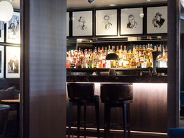 Uma área de bar com diversos tipos de garrafas de bebidas, retratos emoldurados na parede e dois bancos altos.