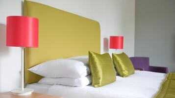 Quarto de hotel com uma cama, roupa de cama branca, almofadas verdes e brancas, uma luminária vermelha em uma mesa de cabeceira e uma luminária vermelha na parede.