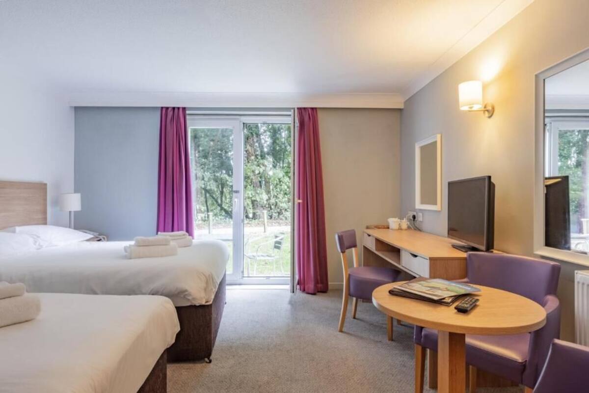 Quarto de hotel com duas camas, uma escrivaninha com cadeira, televisão e janela com cortinas.