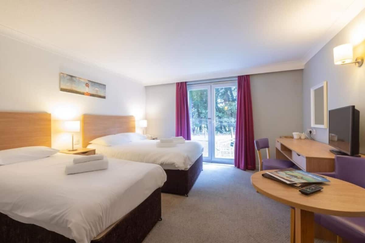 Quarto de hotel com duas camas, uma escrivaninha, televisão e janela com cortinas.