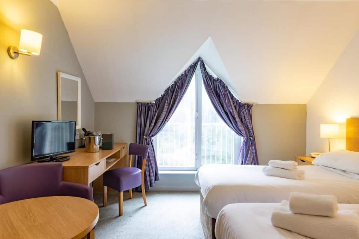 Quarto de hotel com uma cama, uma mesa de trabalho com televisão, uma cadeira e uma janela com cortinas.