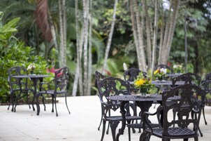 Mesas e cadeiras ao ar livre, com um jardim tropical ao fundo.