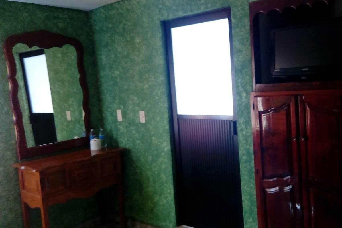 Quarto com paredes verde musgo, um armário de madeira, um espelho, uma porta e uma televisão sobre um suporte de madeira.