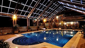 Piscina interna com teto de vidro, deck de madeira e áreas de estar.