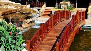 Uma ponte de madeira sobre um espelho d'água, com degraus de pedra e uma pequena construção com plantas.
