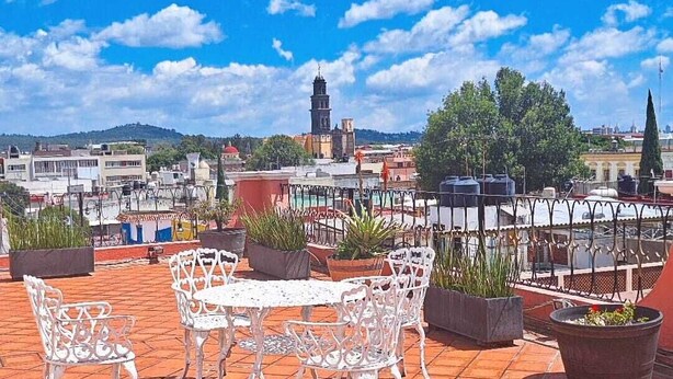 Terraço no telhado com mobiliário de exterior e plantas em vasos, com vista para um horizonte urbano com edifícios e uma torre.