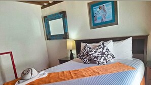 Cama bem arrumada com colcha listrada, duas almofadas estampadas e um cobertor laranja. Há um quadro na parede acima da cama e uma luminária na mesa de cabeceira ao lado.