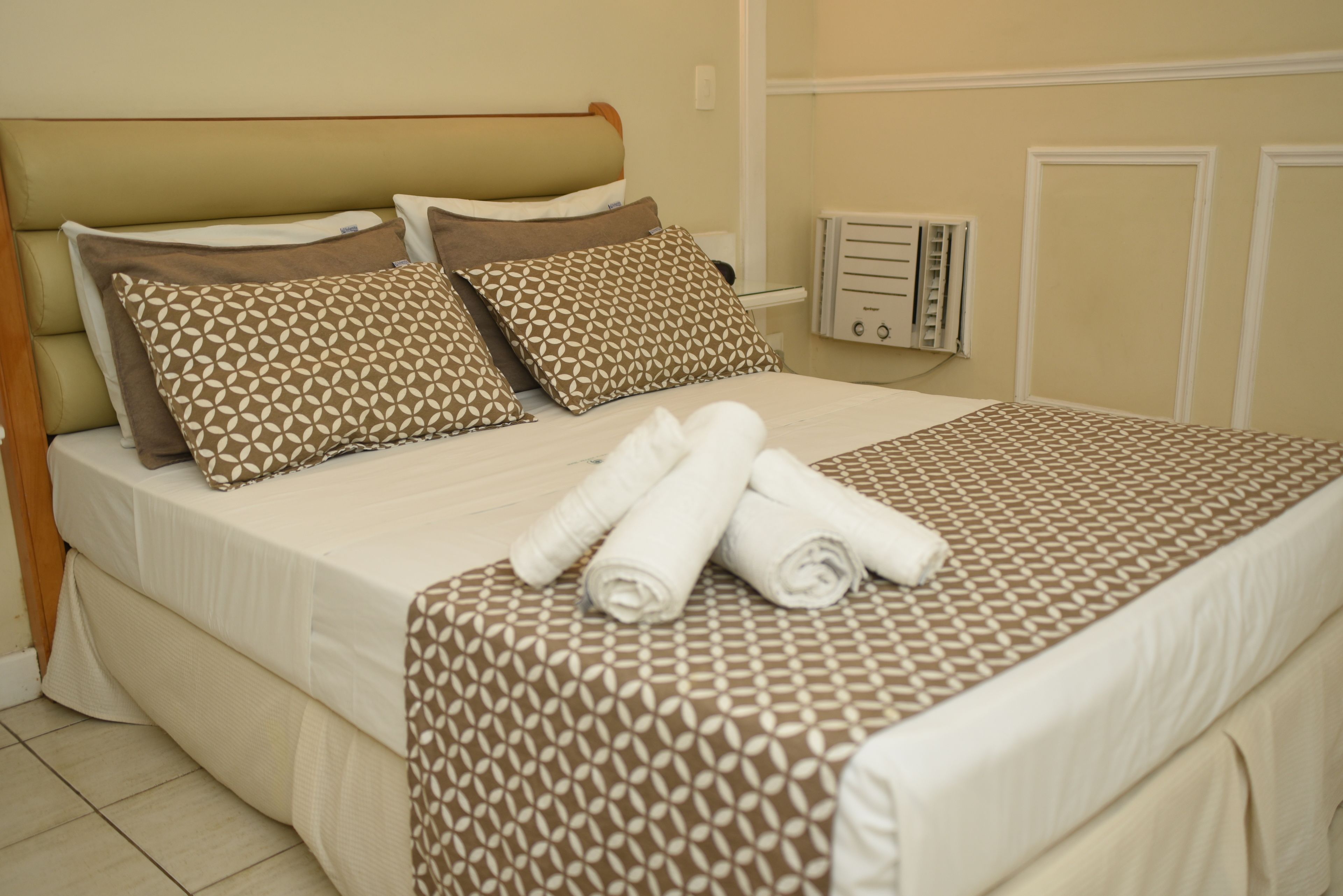 Cama bem arrumada com almofadas estampadas e toalhas enroladas, aquecedor ao lado da cama e parede lisa com uma porta.