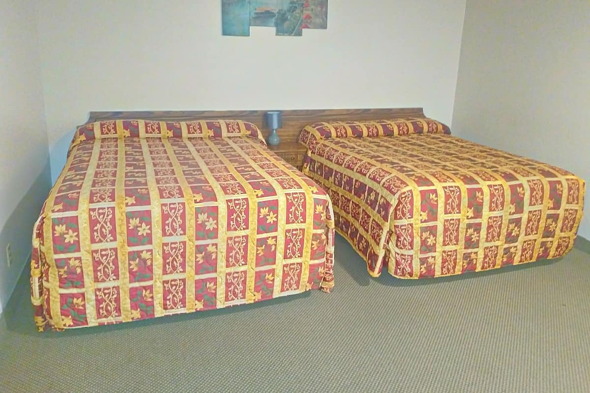 Uma cama de casal com colcha estampada em vermelho e amarelo, cabeceira de madeira e uma pequena luminária azul no criado-mudo.