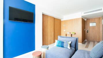 Quarto de hotel moderno com uma parede de destaque azul, uma TV de tela plana na parede, um sofá cinza com almofadas estampadas e um guarda-roupa de madeira.
