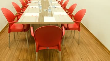Uma sala de conferências com uma mesa comprida, cadeiras vermelhas e garrafas de água.