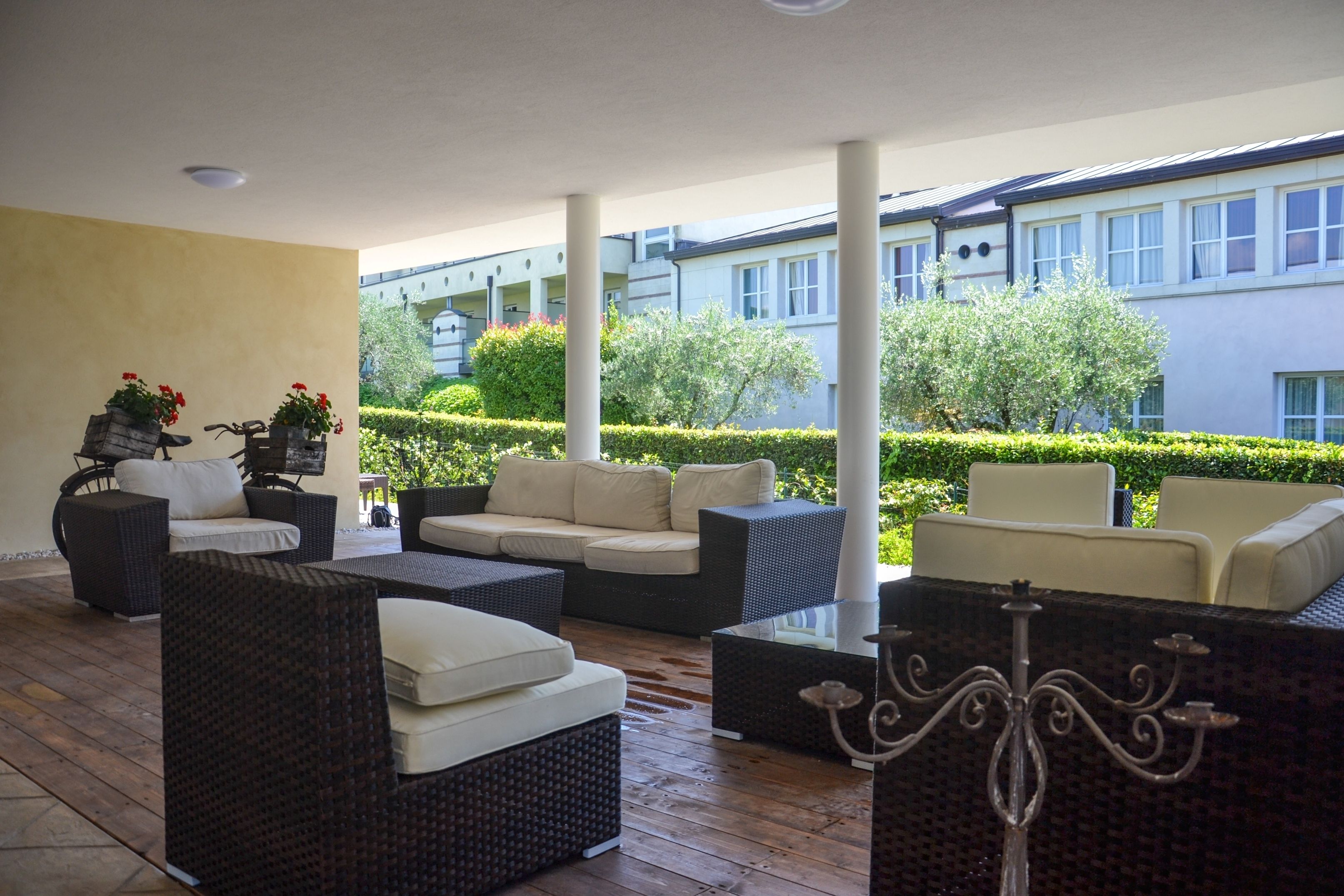Uma área de estar coberta ao ar livre, com mobiliário de vime e uma mesa de vidro, com vista para um jardim bem cuidado e um edifício ao fundo.