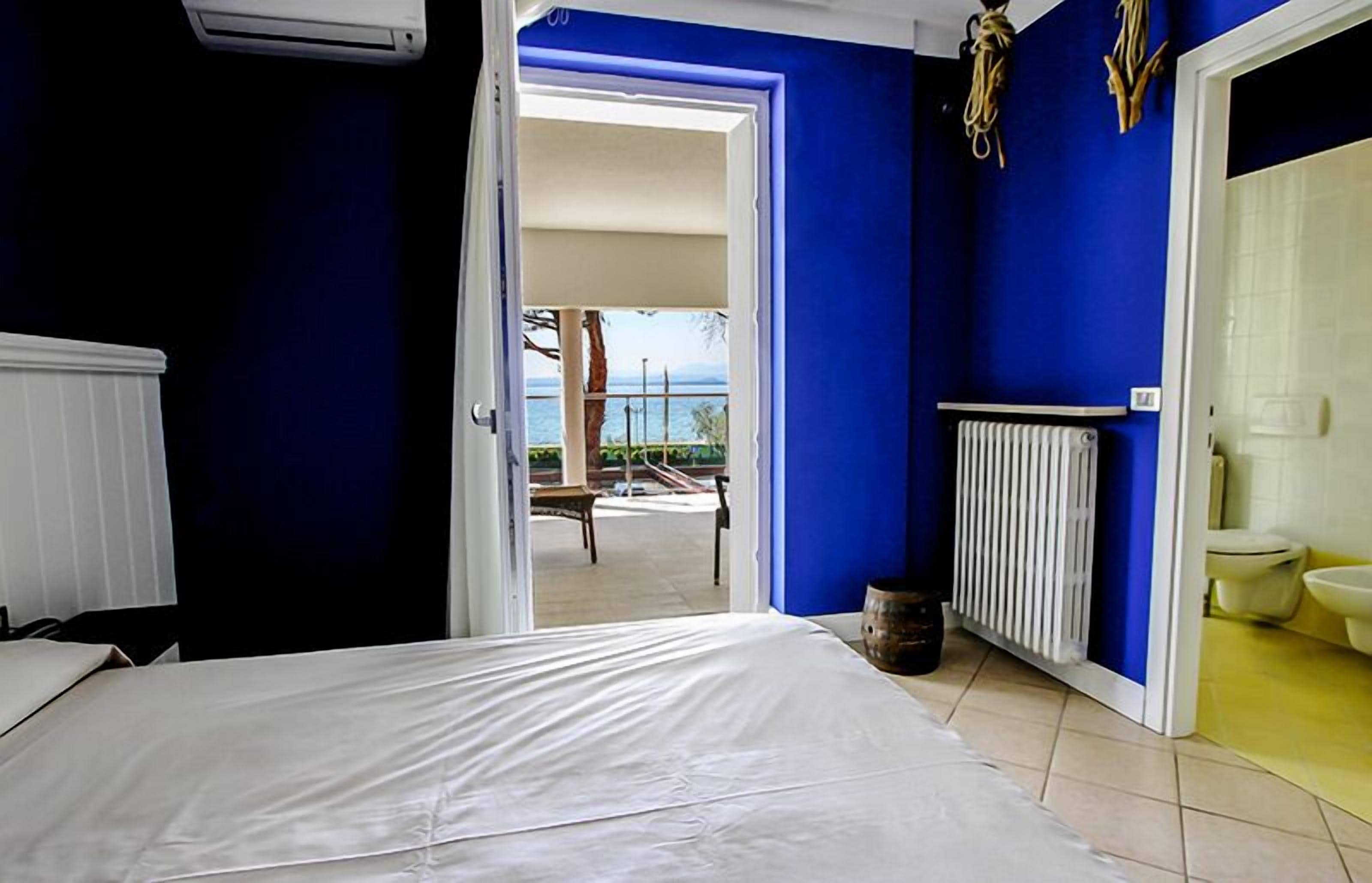 Quarto com cama, radiador, banheiro com vaso sanitário e vista para o mar por uma porta aberta.