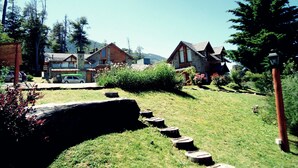 Uma área residencial com casas de pedra e madeira, um caminho de entrada calçado e um muro de pedra com degraus que sobem uma colina gramada.