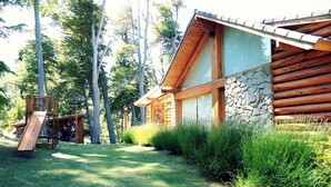 Uma cabana de toras com base de pedra, um deck de madeira e um gramado cercado por árvores altas.