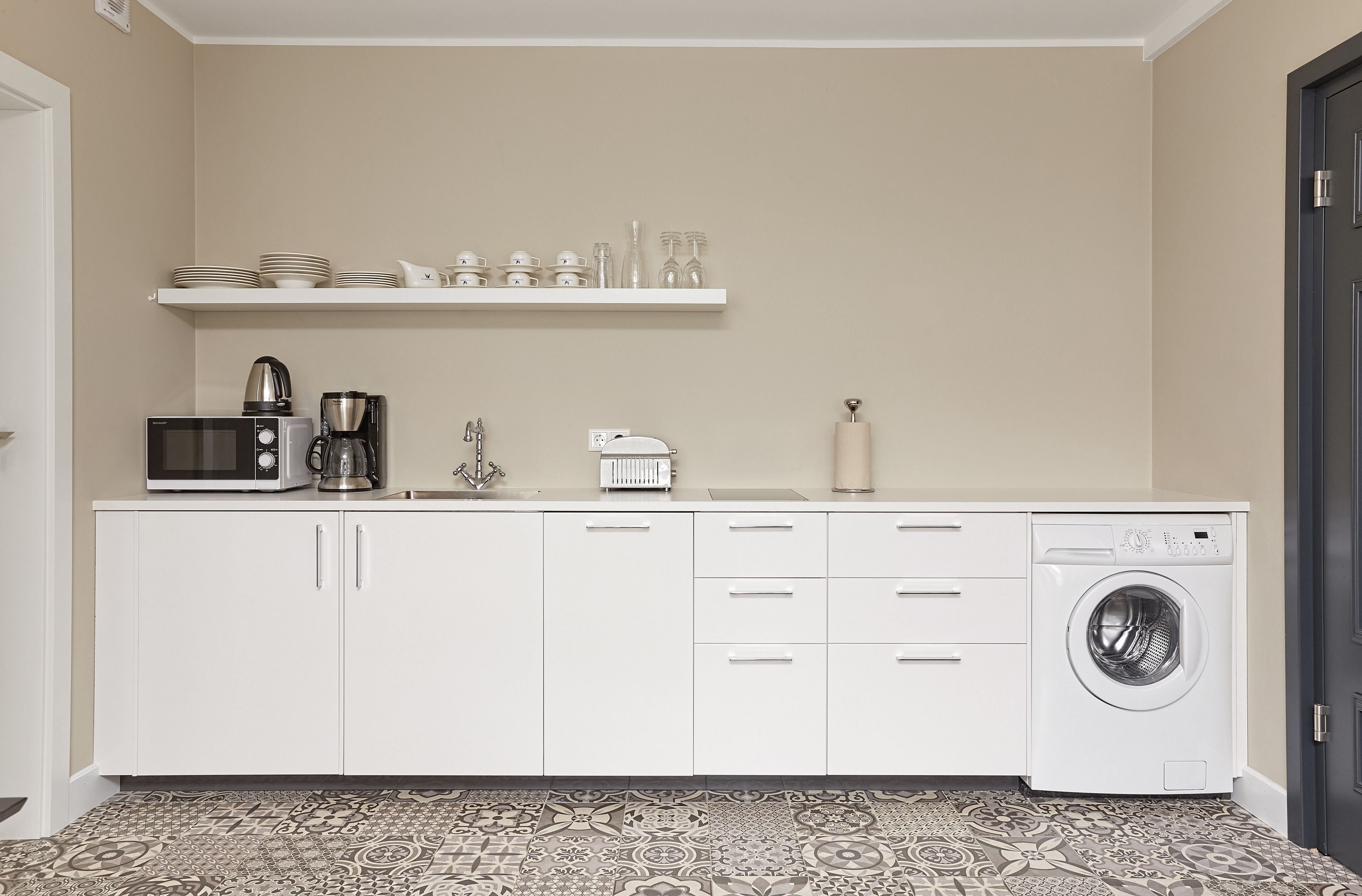 Uma cozinha compacta com armários brancos, micro-ondas, máquina de café, torradeira, máquina de lavar e piso estampado.