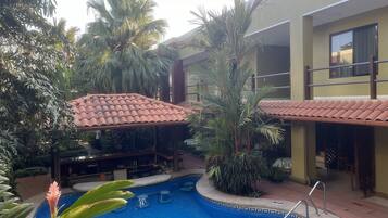 Piscina com deck ladrilhado, rodeada por palmeiras e uma área de estar coberta.