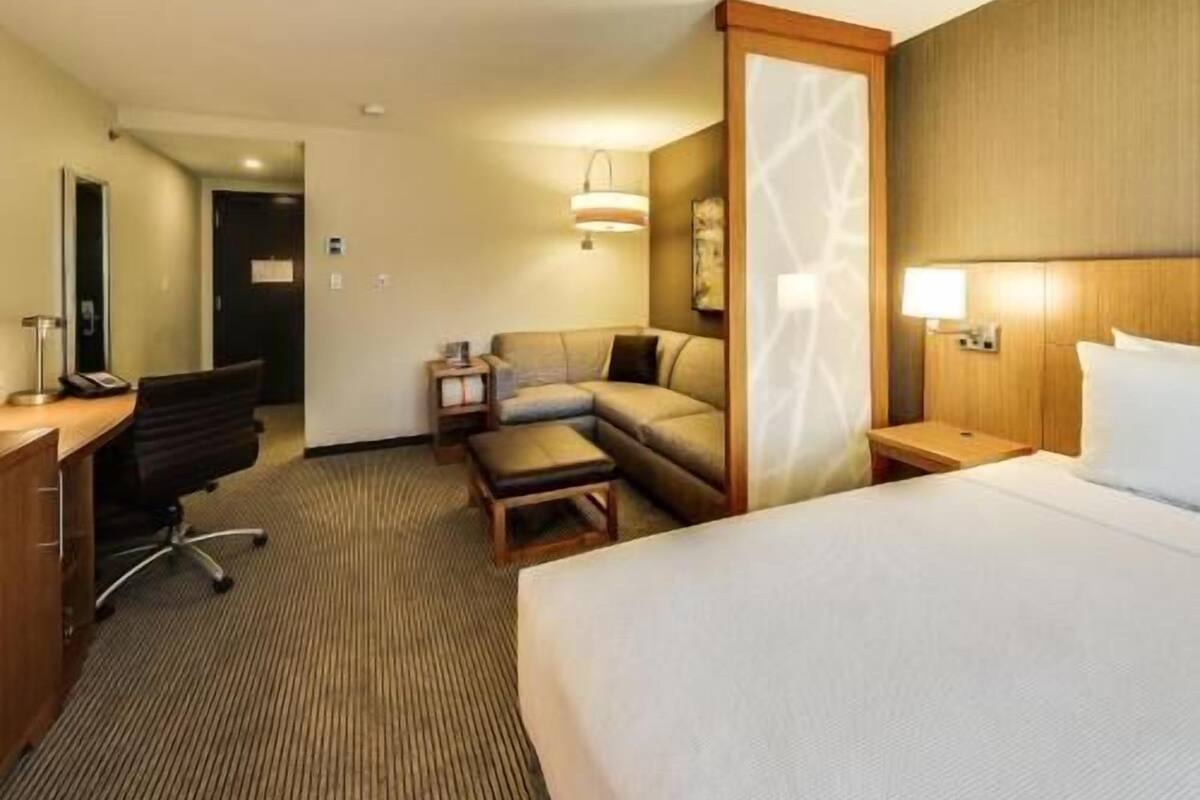Quarto de hotel com cama, escrivaninha, cadeira, sofá, mesa pequena e mesa de cabeceira.