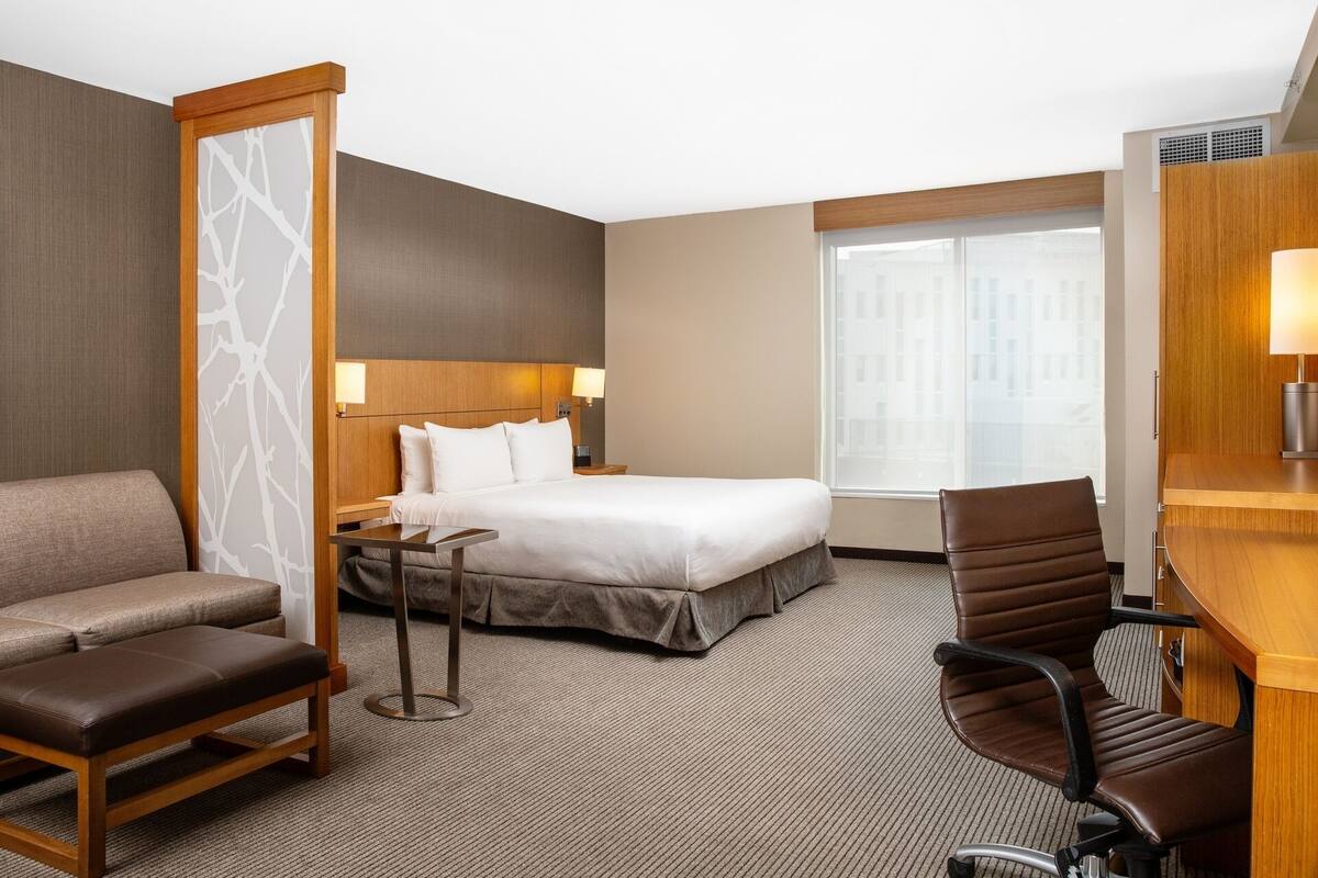 Quarto de hotel com cama, escrivaninha, cadeira, sofá e uma grande janela.
