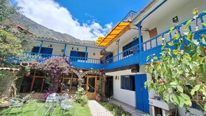 Um edifício de dois andares com paredes externas azuis e brancas, uma varanda com toldo amarelo e um jardim com plantas em vasos e área de estar ao ar livre.