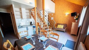 Uma área de refeições com uma mesa de madeira posta para dois, uma escada, uma cozinha e uma sala de estar com sofá e televisão.