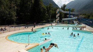 Uma grande piscina ao ar livre cercada por árvores e montanhas, com pessoas nadando e brincando.