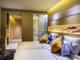 Quarto de hotel moderno com duas camas, uma janela grande com persianas, uma obra de arte na parede e uma porta de madeira.