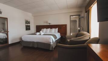 Quarto de hotel com cama, cadeira, escrivaninha, espelho e pintura na parede.