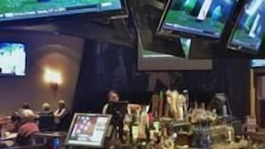 Uma área de bar com várias telas exibindo conteúdo, um balcão com diversos tipos de bebidas e piso coberto por carpete.
