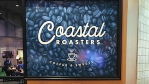 Uma cafeteria Coastal Roasters com um grande letreiro exibindo grãos de café, duas cadeiras de madeira e um suporte de menu.
