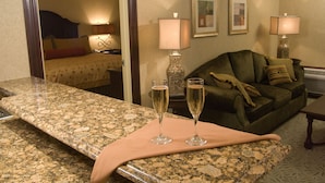 Quarto de hotel com um espelho grande, uma cama com roupa de cama florida, um sofá verde, duas luminárias de chão e duas taças de champanhe sobre uma mesa de mármore.