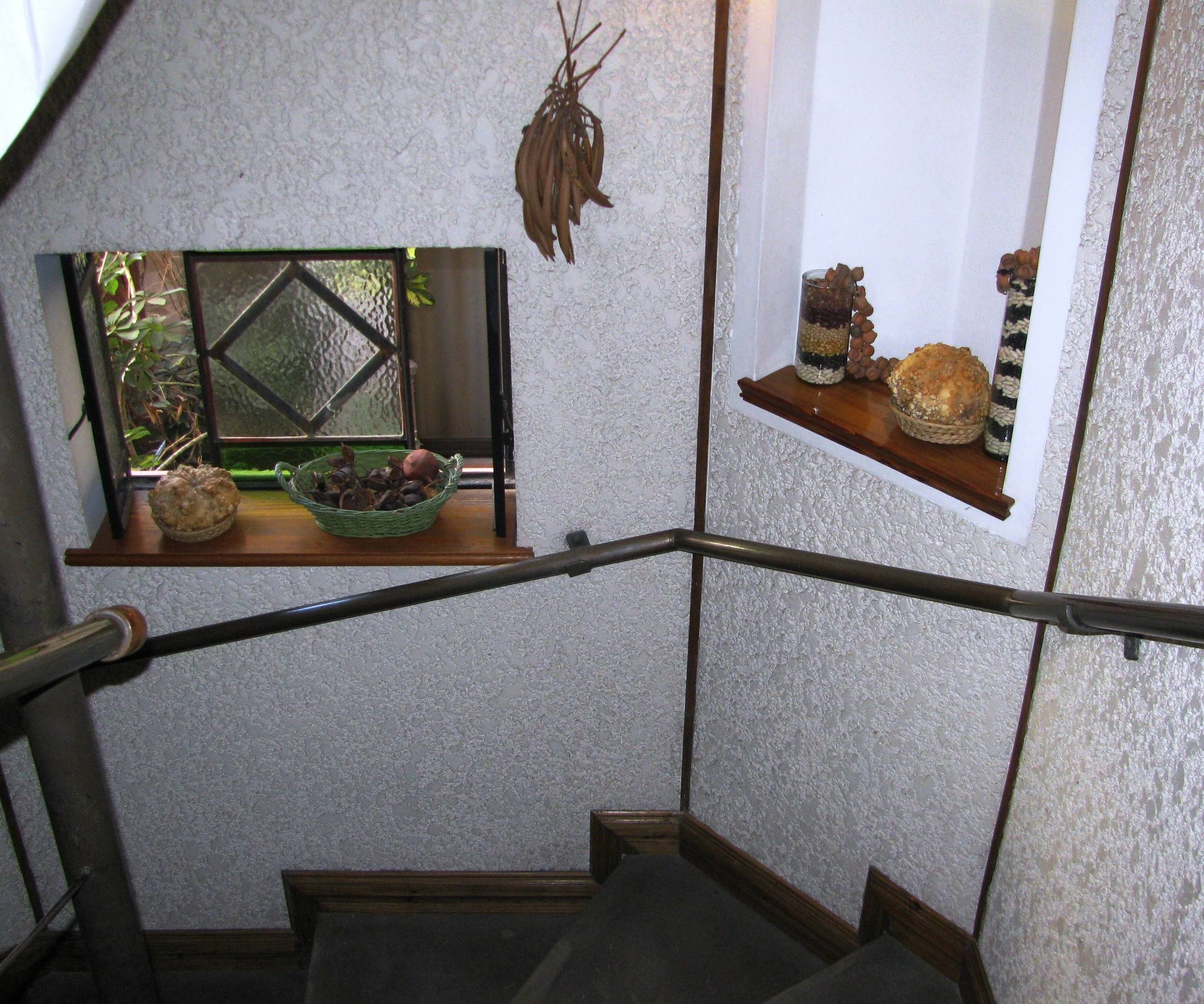 Uma escada com corrimão, uma janela com vista para o verde e uma prateleira com produtos de panificação.