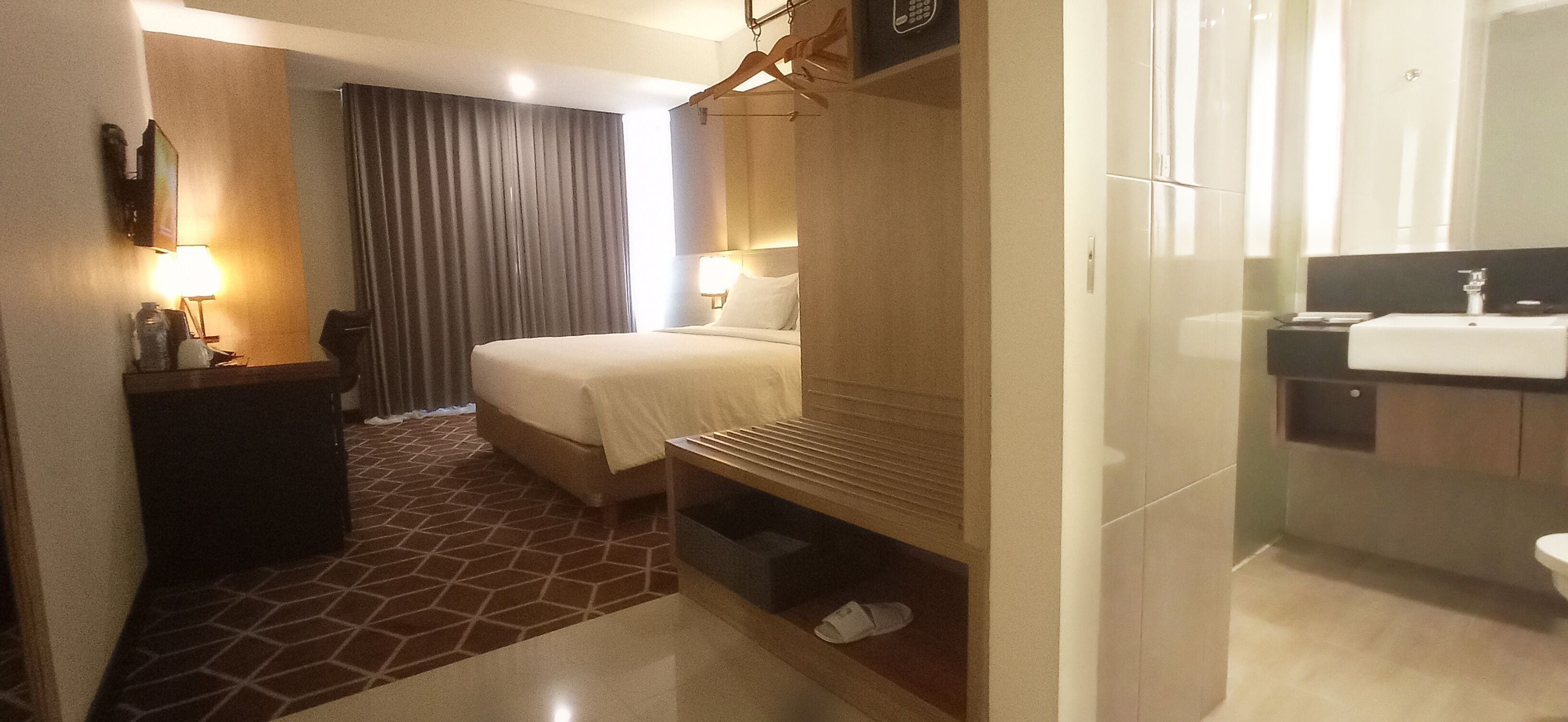Quarto de hotel com cama, mesa de cabeceira, luminária e banheiro com pia e espelho.