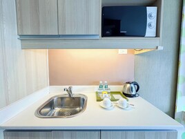 Uma cozinha compacta com uma pia de aço inoxidável, micro-ondas e chaleira sobre uma bancada branca.