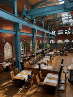 Um restaurante espaçoso com paredes de tijolo, mesas de madeira e vigas azuis estruturais.