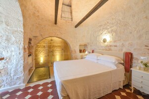 Um quarto com parede de pedra, uma cama, uma mesinha de cabeceira e uma janela pequena.