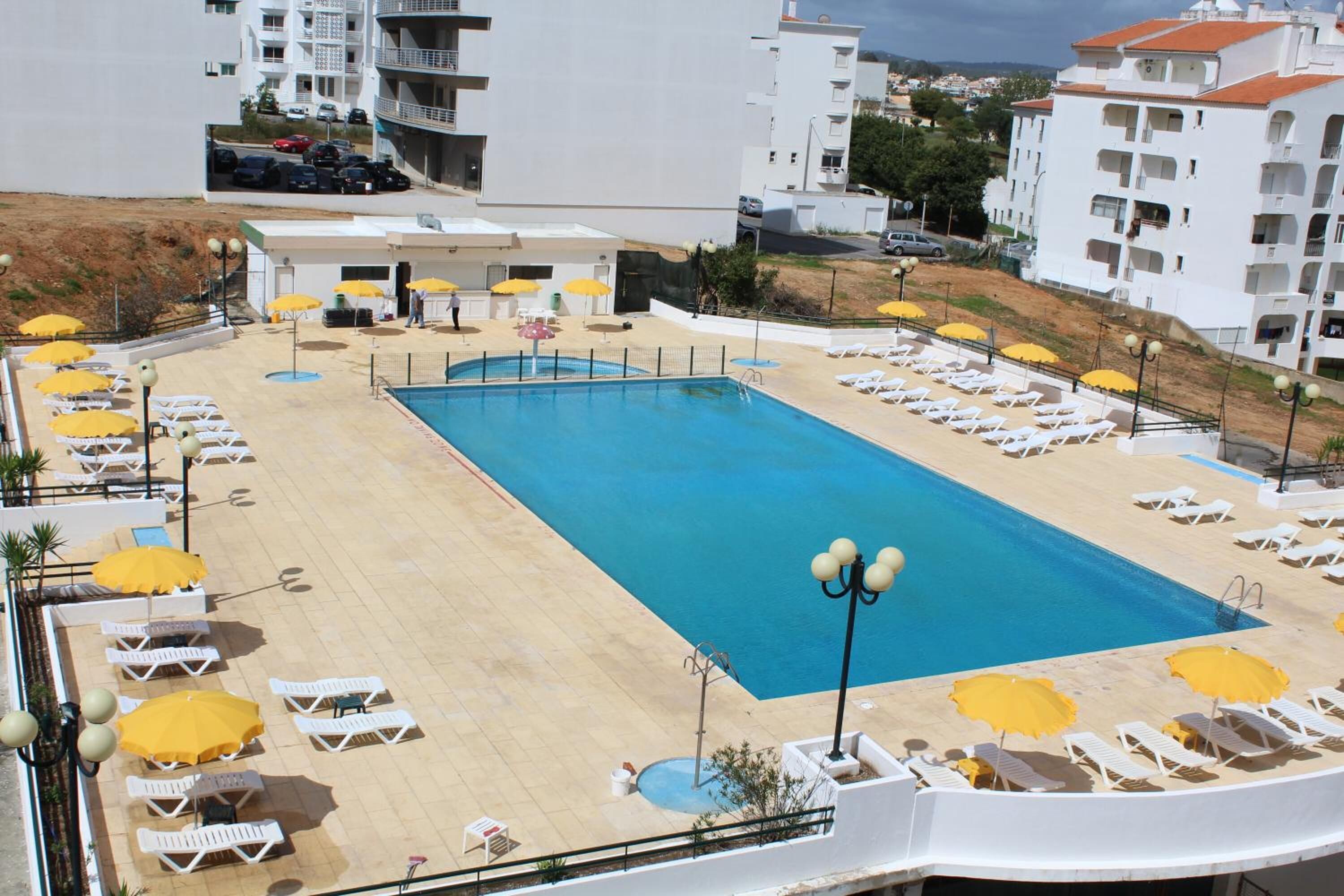 Uma grande piscina ao ar livre rodeada por espreguiçadeiras e guarda-sóis, com edifícios ao fundo.