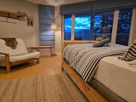 Quarto com cama, cadeira, mesa de cabeceira, luminária e janela com vista para árvores.