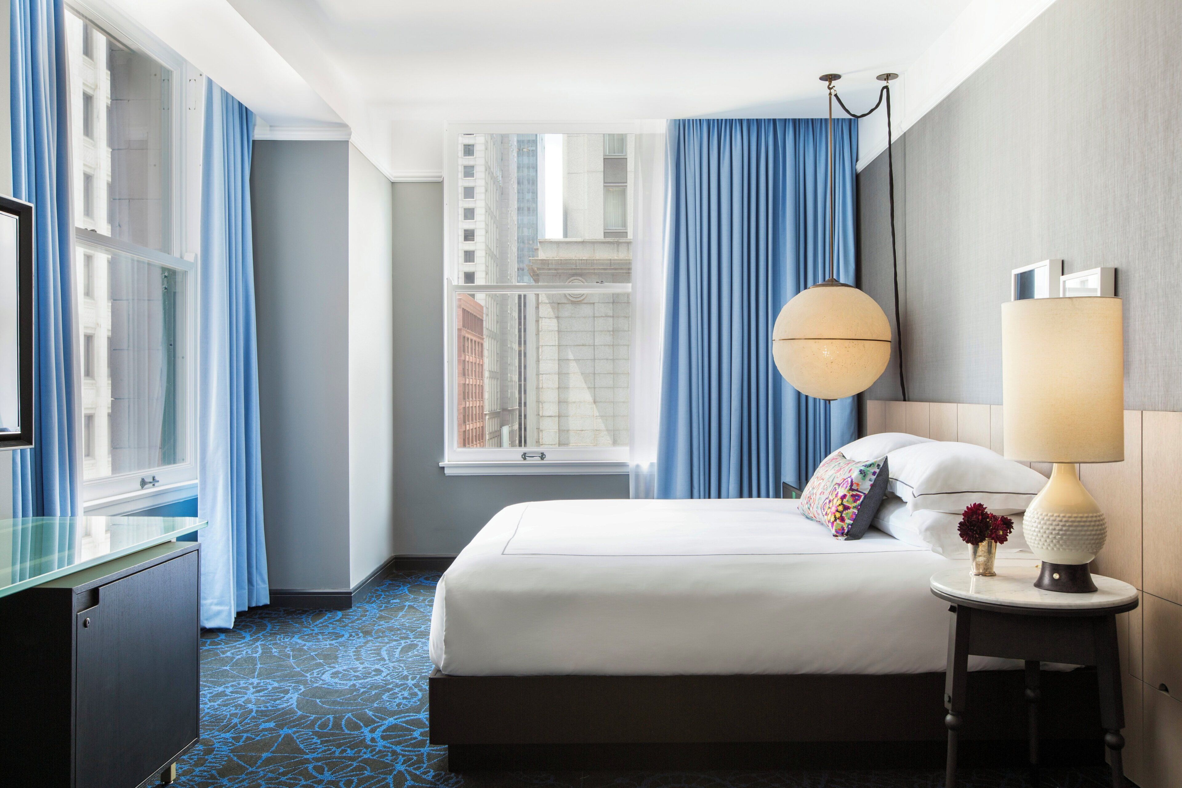 Quarto de hotel com cama, mesa de cabeceira com luminária, janela com cortinas e vista para a cidade.