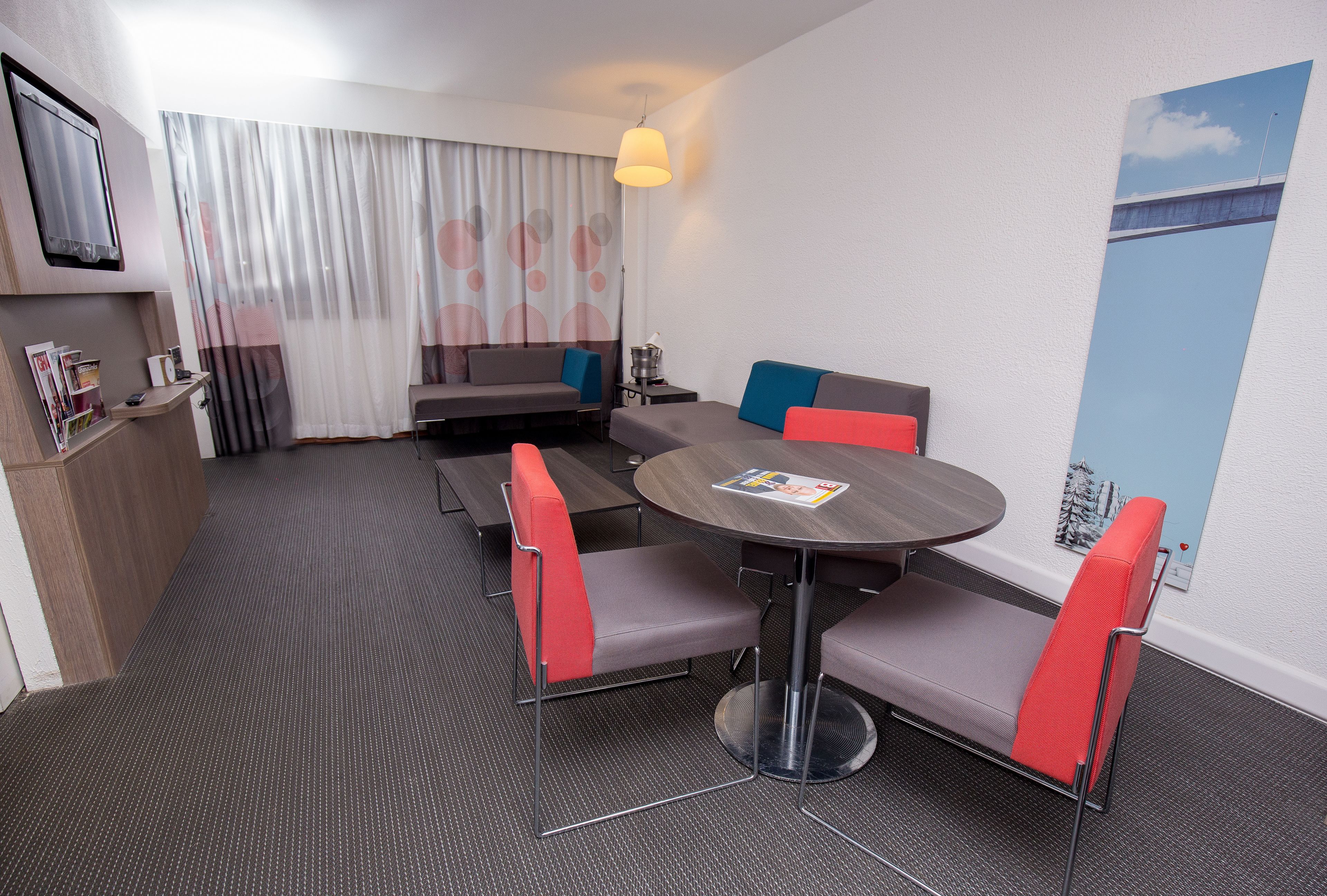 Quarto de hotel moderno com uma mesa redonda, cadeiras vermelhas e cinzentas, um sofá e uma janela grande com cortinas.