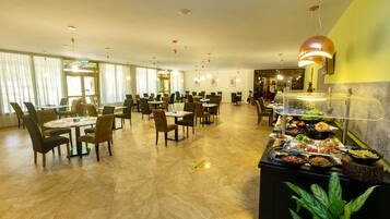 Uma sala de jantar ampla com várias mesas e cadeiras, um buffet com diversos pratos e um lustre moderno.