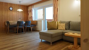 Uma sala de estar acolhedora com piso de madeira, um sofá cinzento, uma mesa de jantar com cadeiras e uma janela com cortinas.