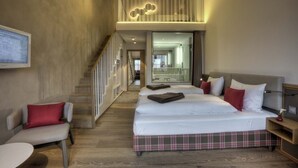 Quarto de hotel com uma cama grande, uma cadeira, uma escrivaninha e uma escada que leva a um andar superior.